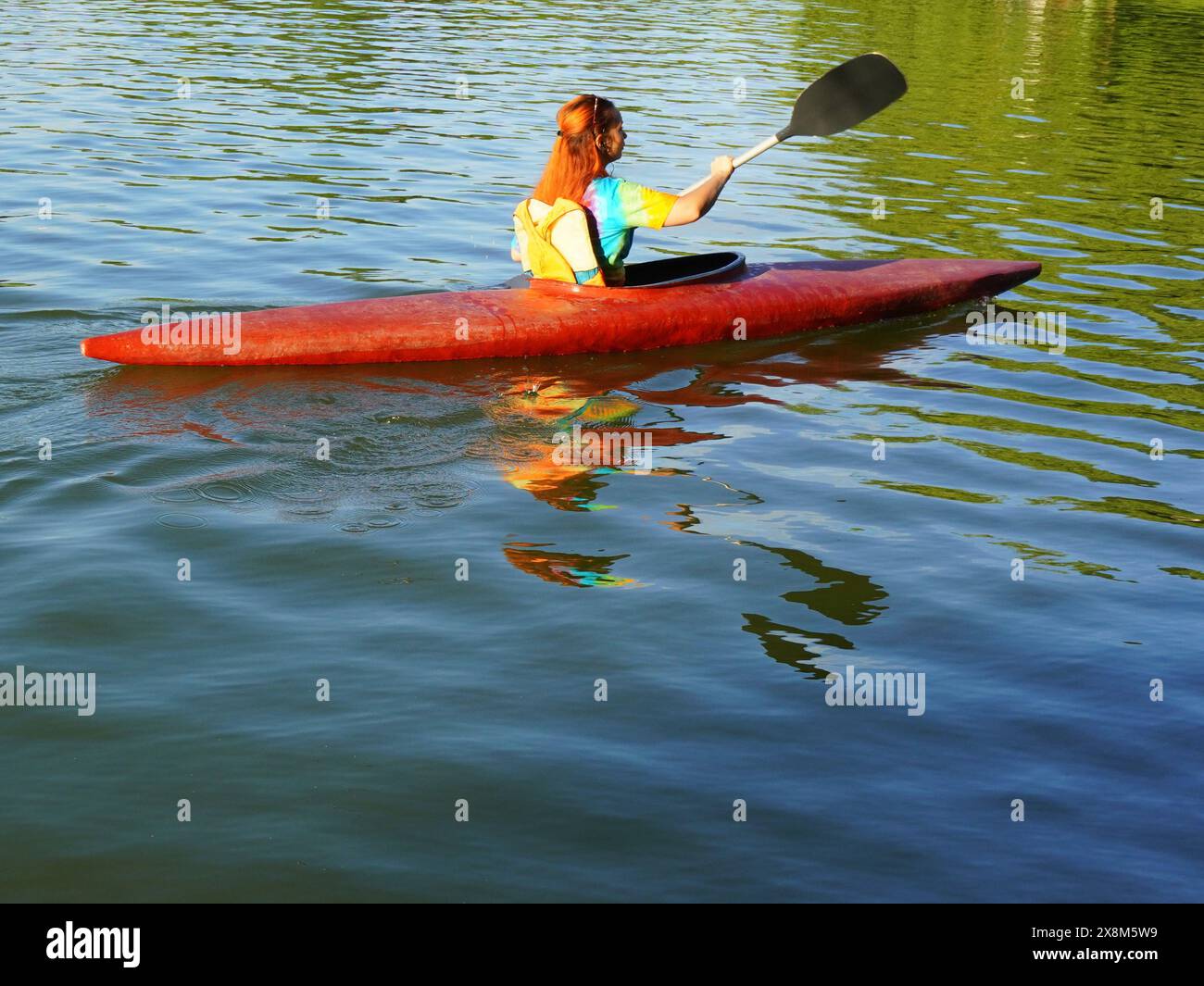 Blonde young woman wearing bright colors, paddling in a kayak on lake ...