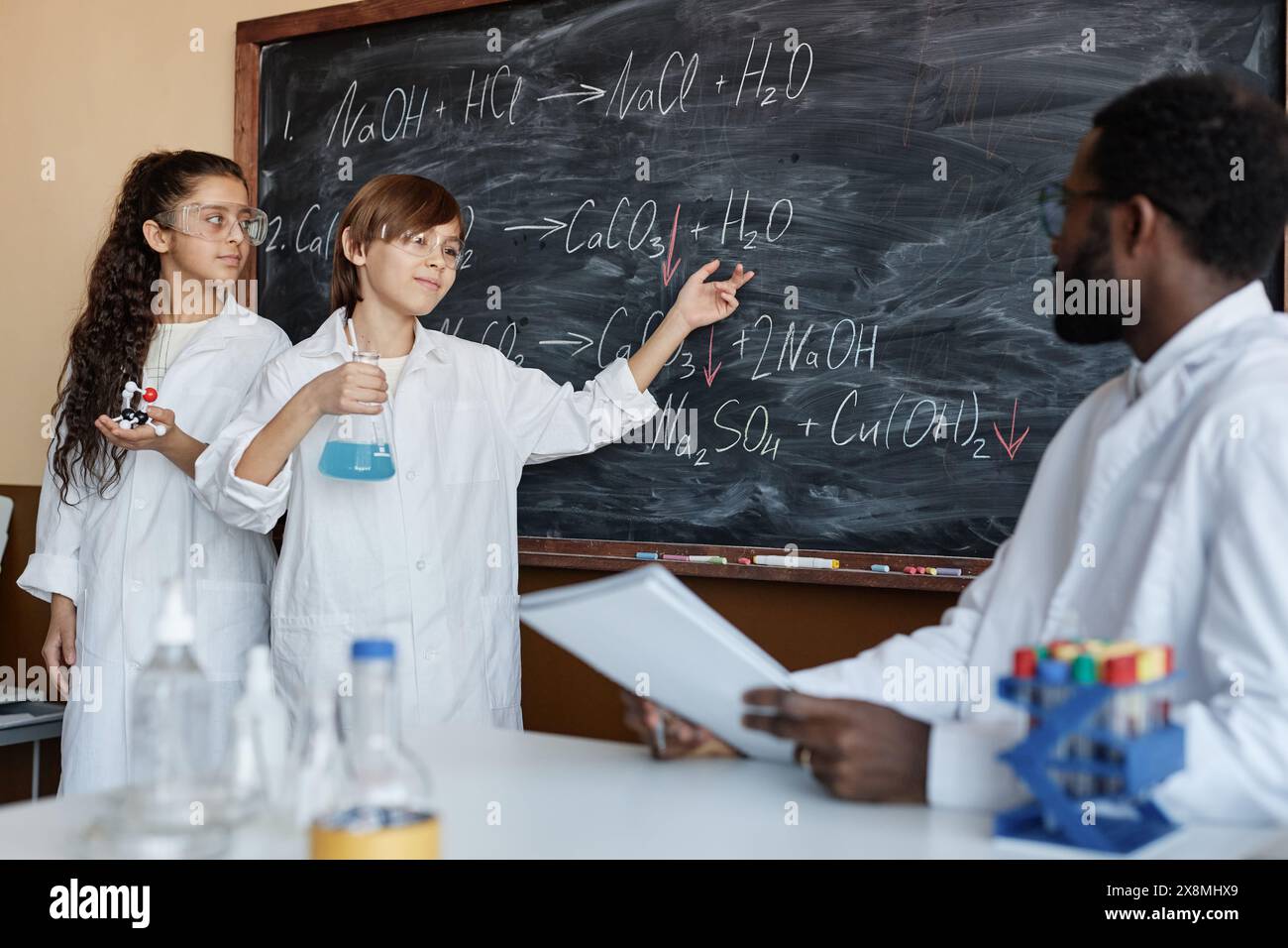 Two diverse students wearing laboratory coats standing at blackboard in classroom solving ...
