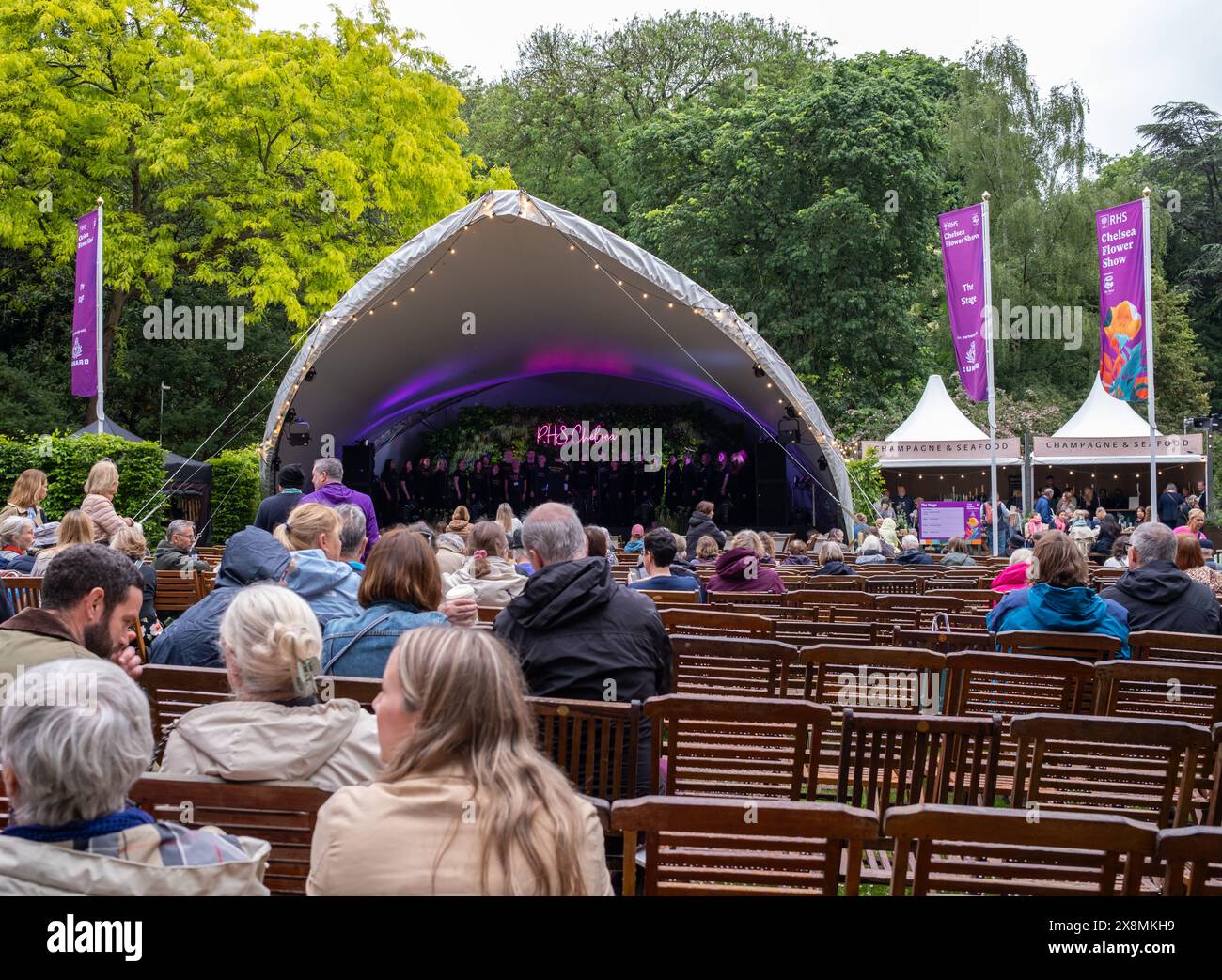 Attendees sitting on wooden chairs watching a choir on the main stage ...