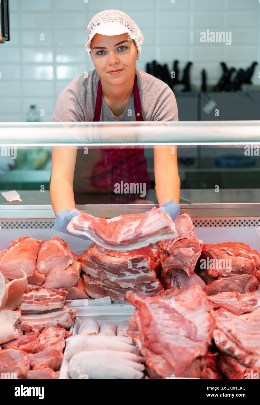 Young saleswoman of butcher shop laying out raw pork ribs in display ...