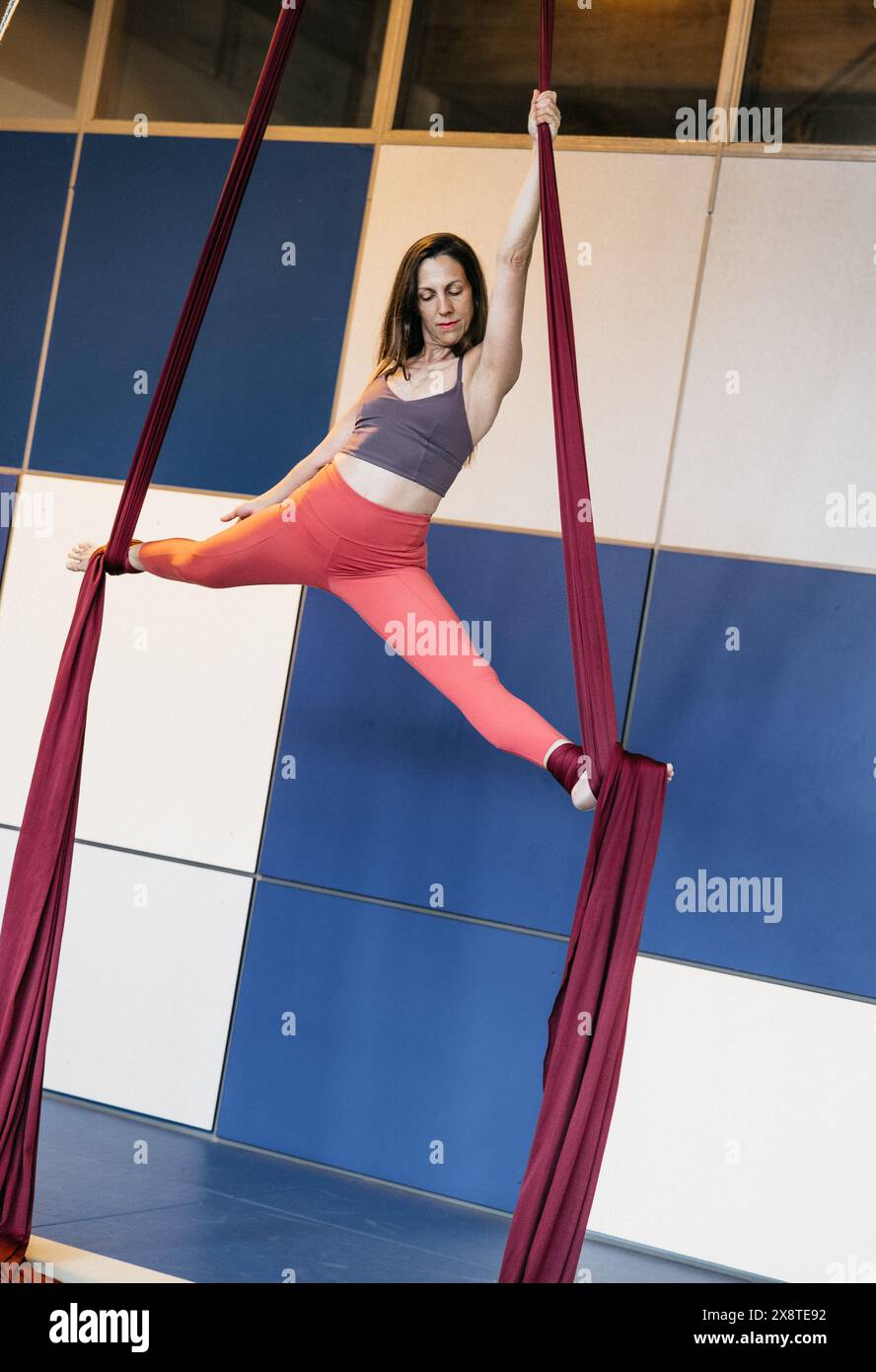 A woman performs an acrobatic pose while hanging from maroon aerial ...