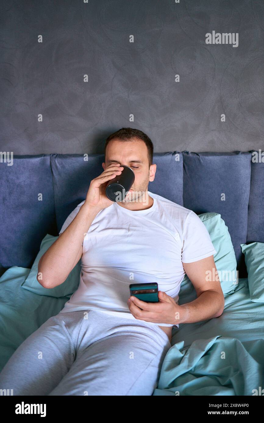 young man drinking morning coffee in bed and scrolling in smartphone ...