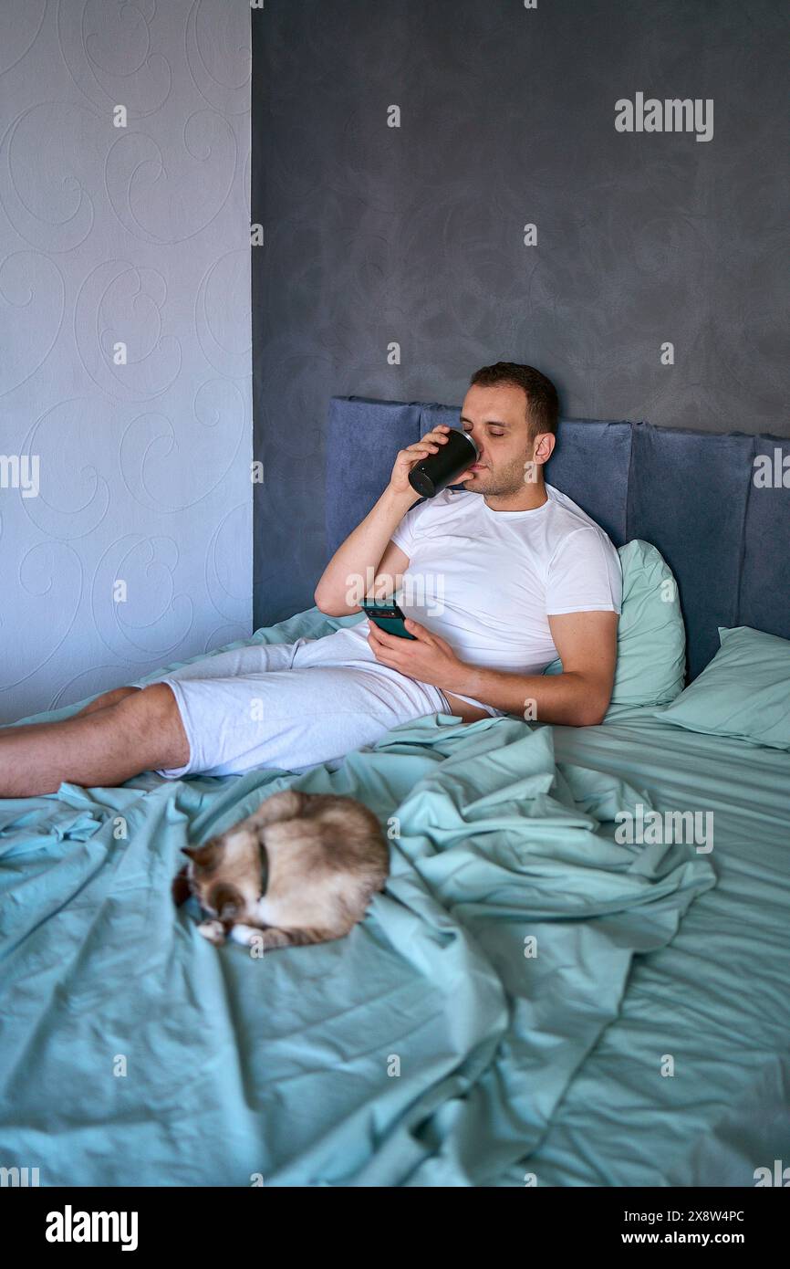 young man drinking morning coffee in bed and scrolling in smartphone ...