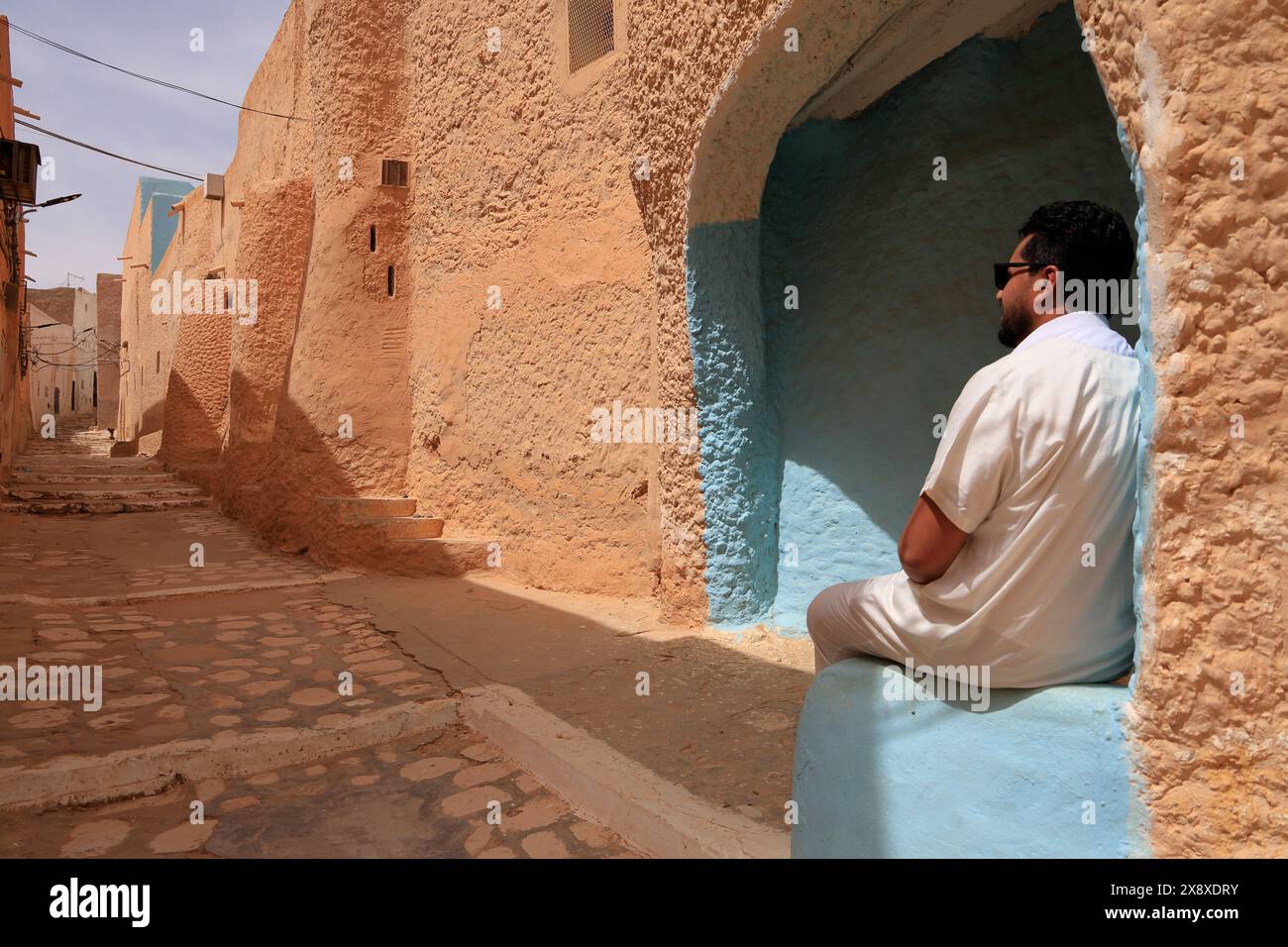 A man in a white robe in street of Ksar Beni Isguen with traditional architectures beside him ...