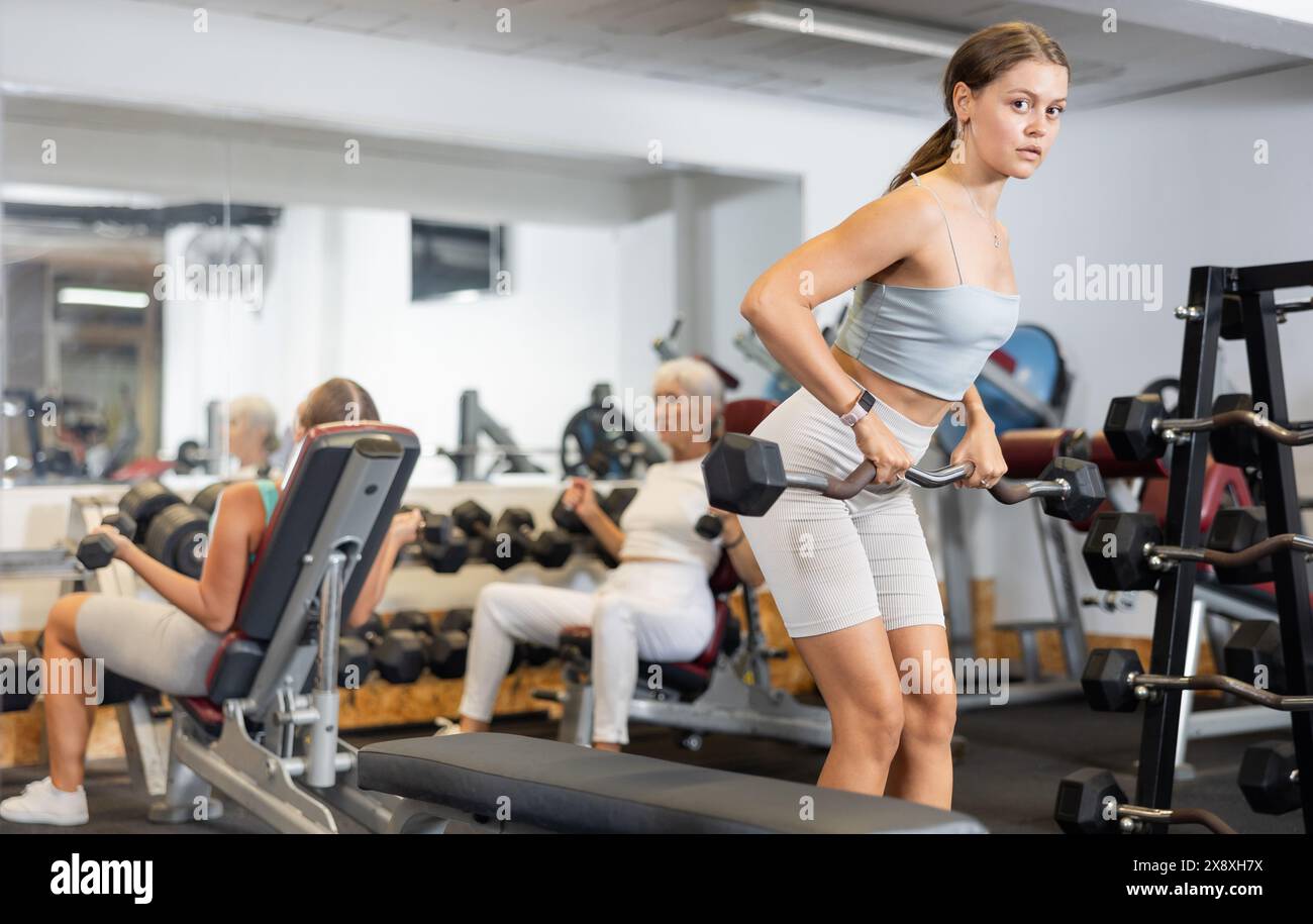 Slender girl trains muscles of arms and back using barbell and ...