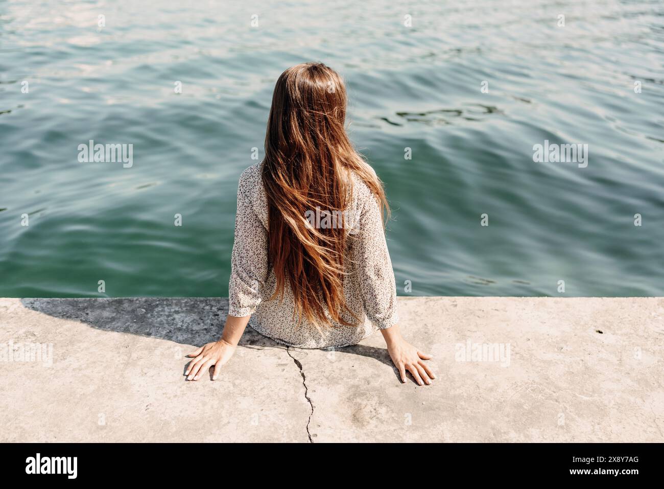 A woman is sitting on a ledge by a body of water, wearing a dress with ...