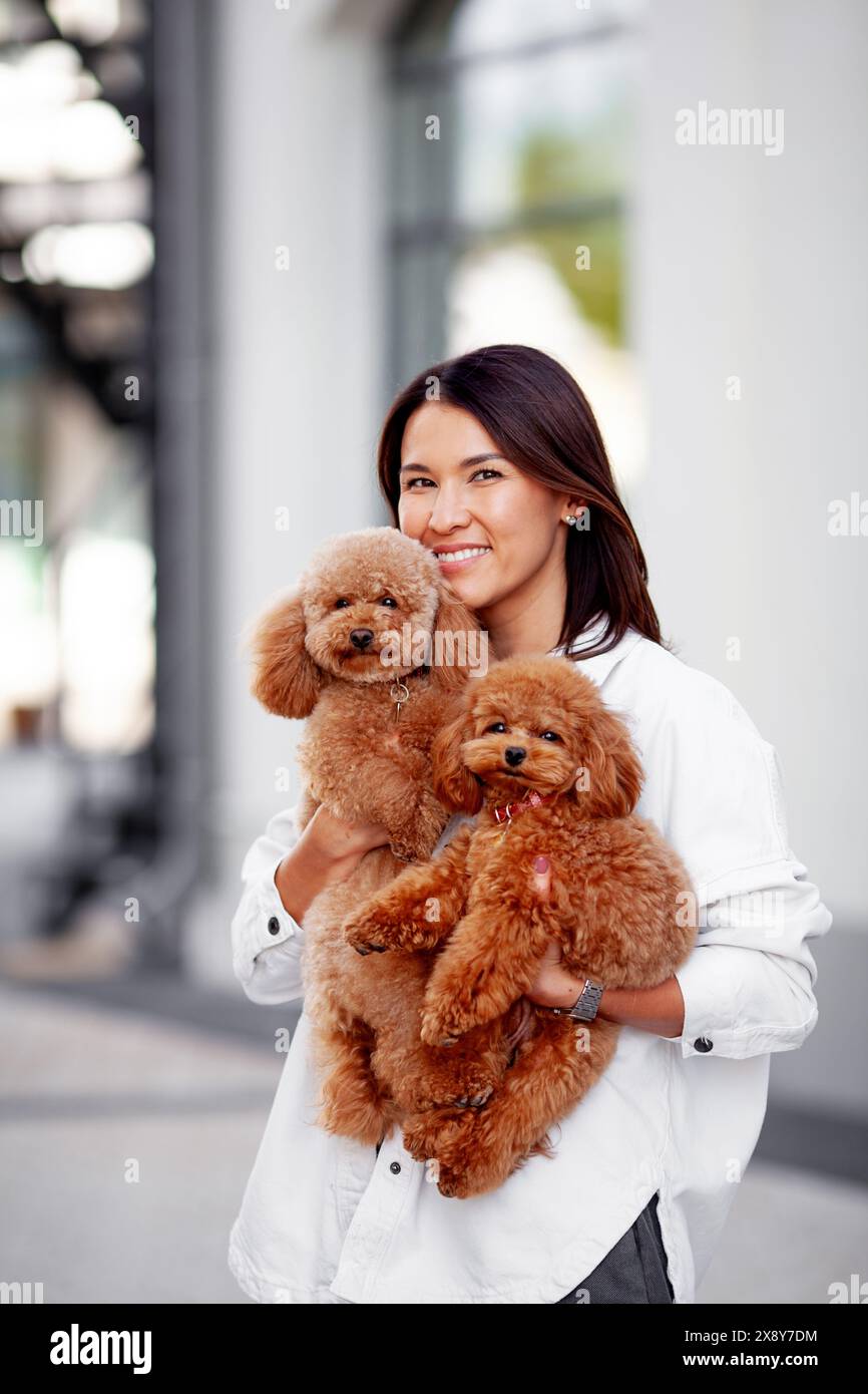 Cheerful woman holds two adorable dogs poodles, smiling while standing ...