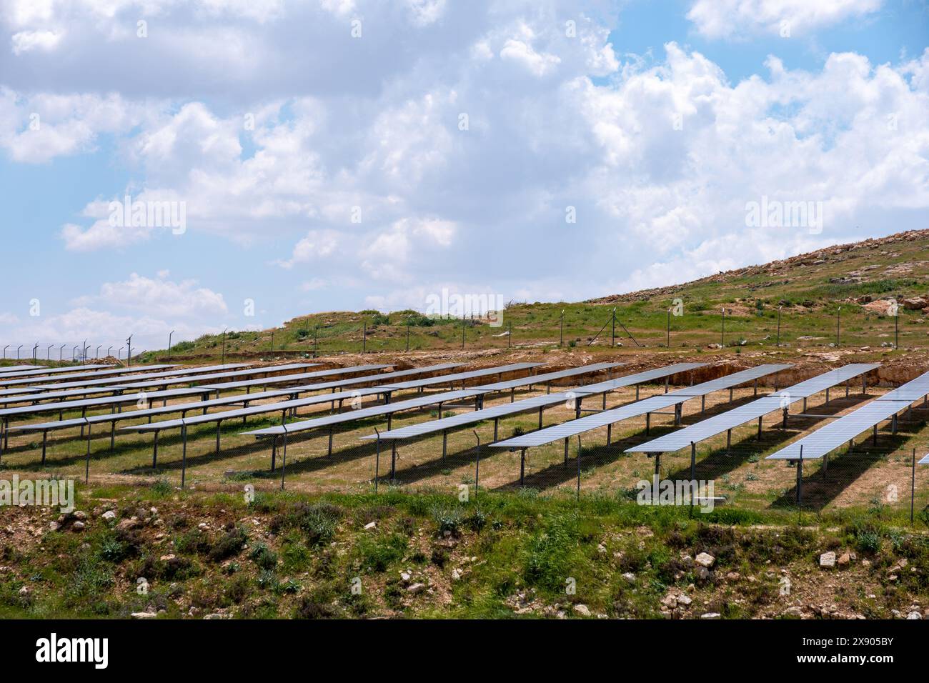 A solar farm is built on a hill under a cloudy sky, surrounded by ...