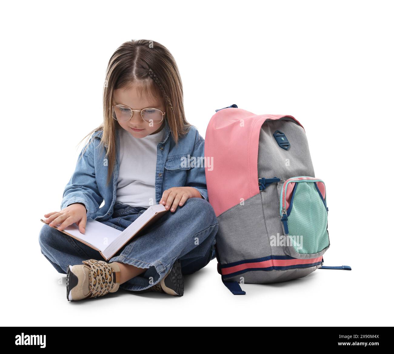 Cute little girl reading book near backpack on white background Stock ...