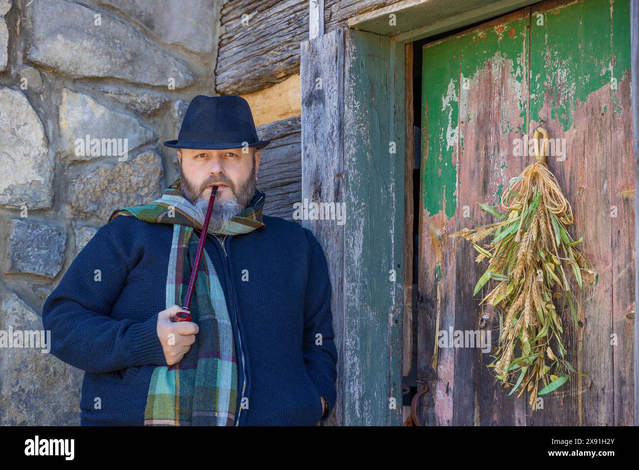 Wearing a hat, plaid scarf and holding a pipe a man leans up against an ...