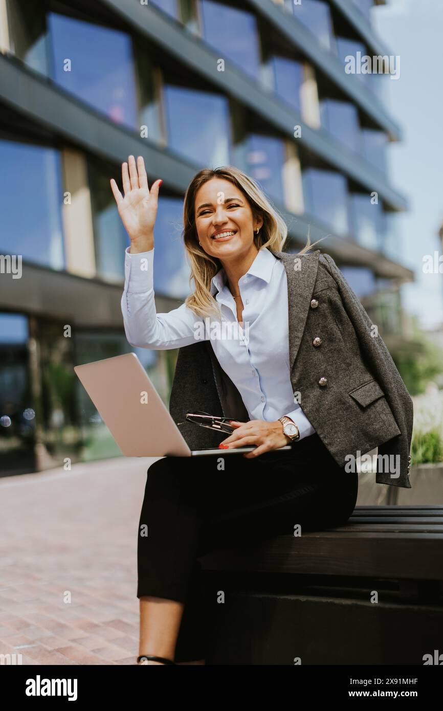Cheerful professional woman with a laptop waves hello while sitting on ...