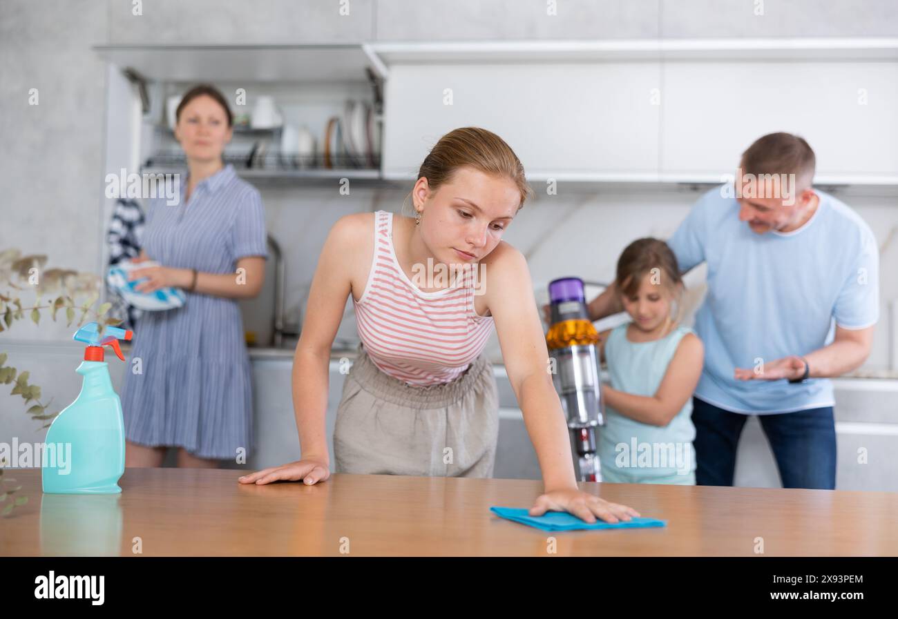 Teen girl cleans table with rag, family clean kitchen in background ...