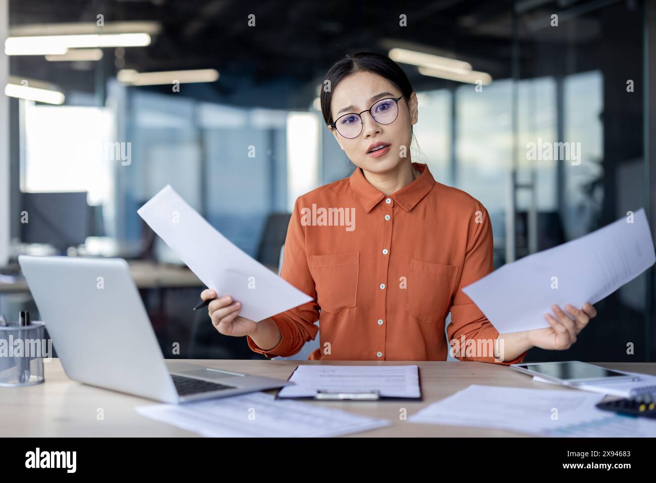 Confused businesswoman wearing glasses reviews documents at her desk in ...