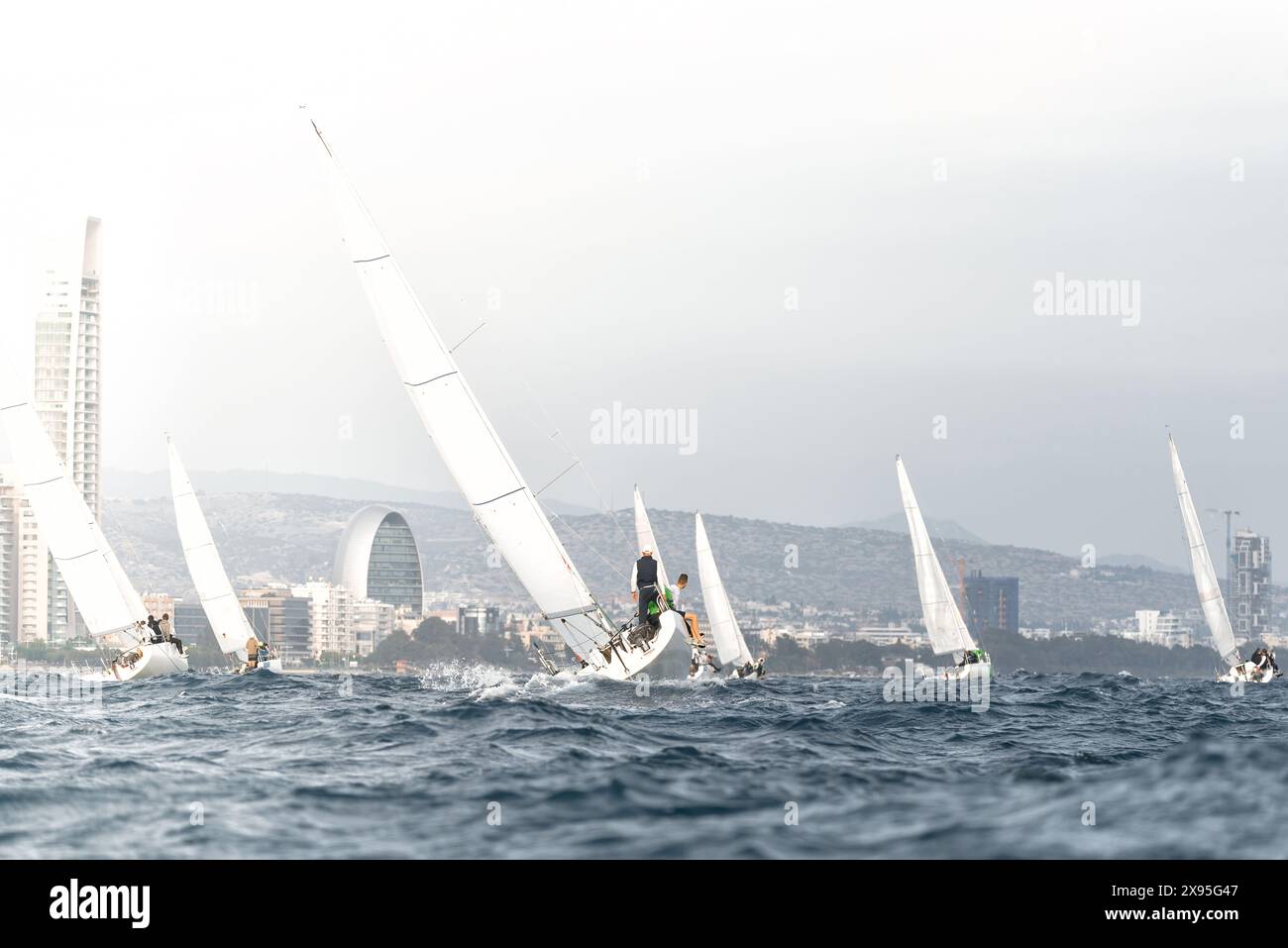 Competitive sailboats race on choppy waters with a coastal city skyline ...