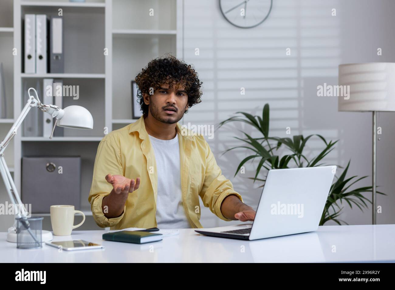 Confused man in yellow shirt sitting at desk with laptop, expressing ...