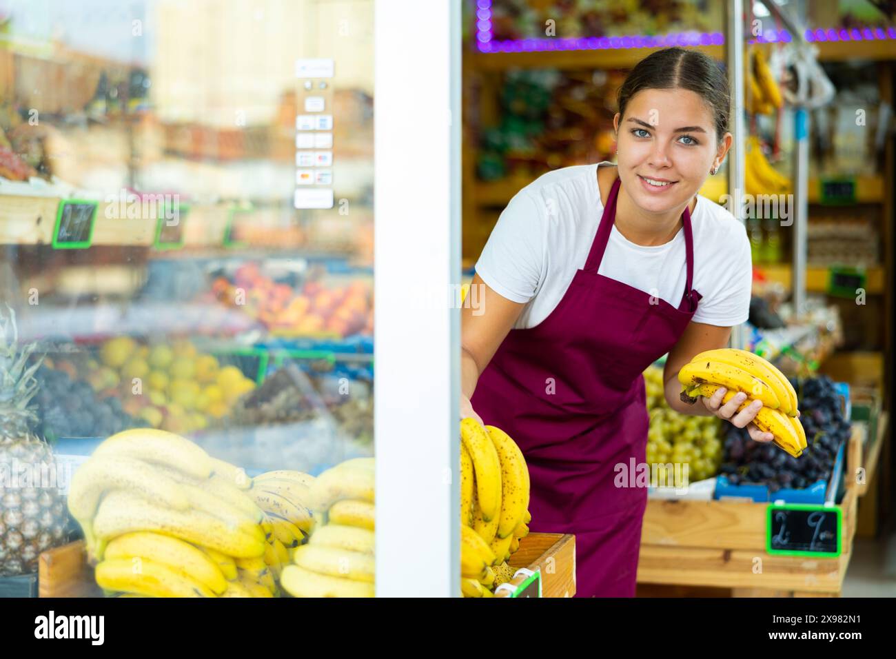 Smiling female merchandiser in uniform laying out bananas at grocery ...