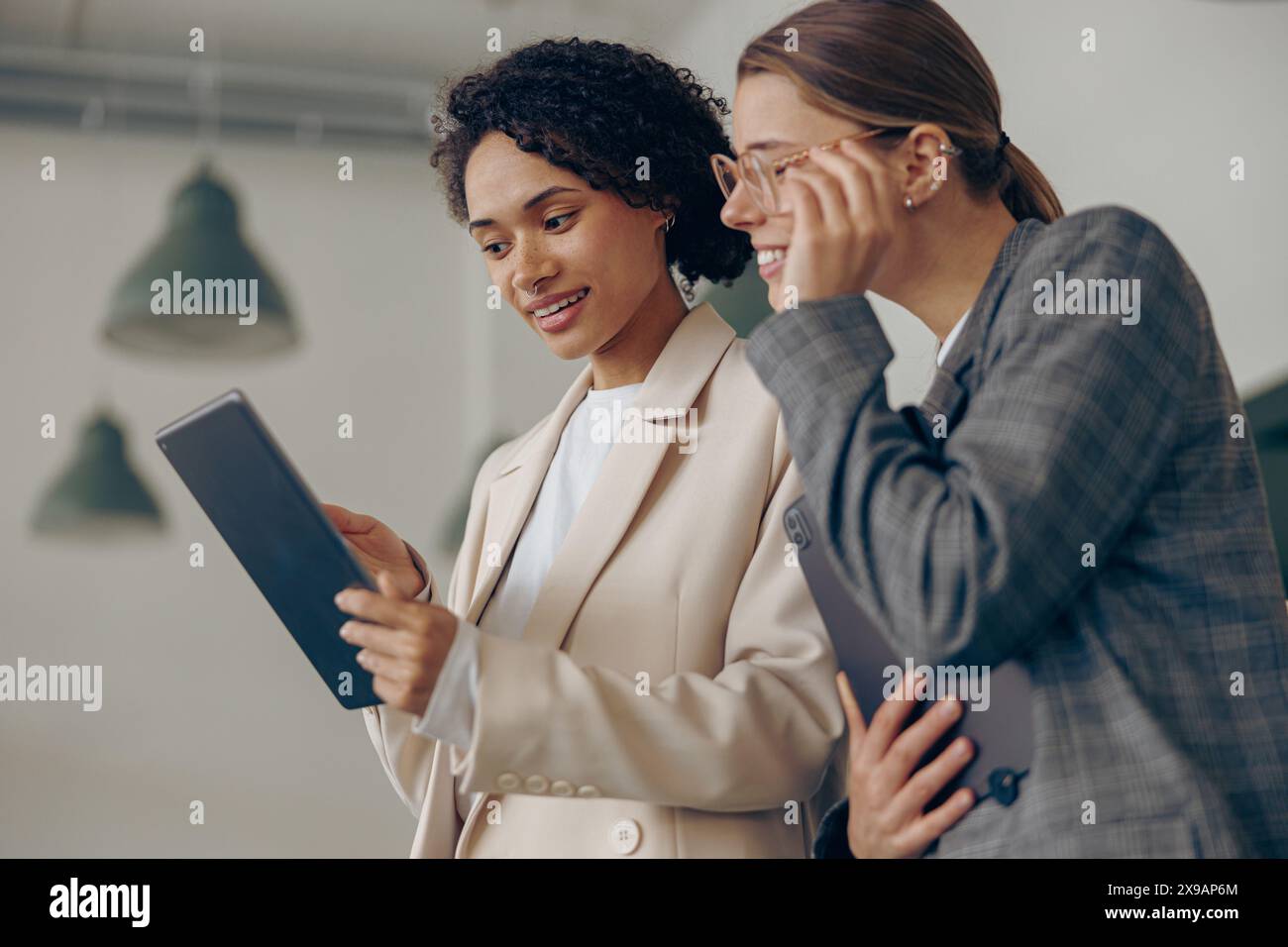 Two busy female coworkers discussing project standing in modern office and use digital tablet ...