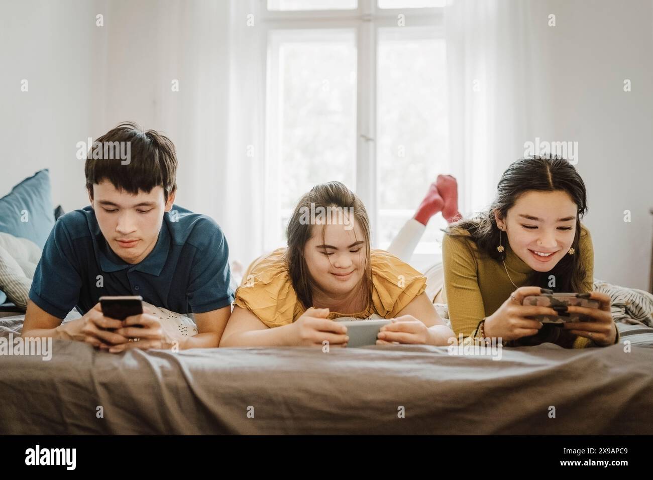 Smiling siblings using smart phones while lying down on bed with down ...