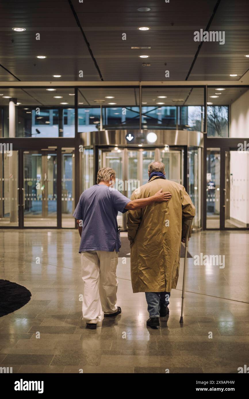 Rear view of doctor walking with senior patient in lobby of hospital ...