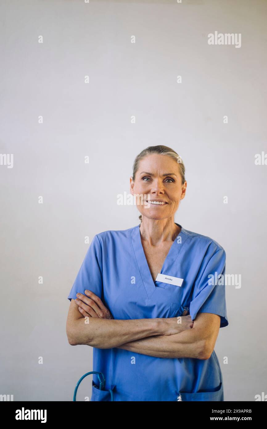 Portrait of smiling senior female doctor standing with arms crossed in front of white wall Stock ...