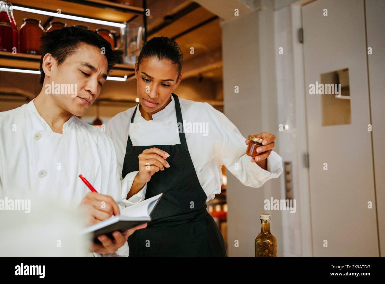 Focused male chef writing with female chef holding jar while standing ...