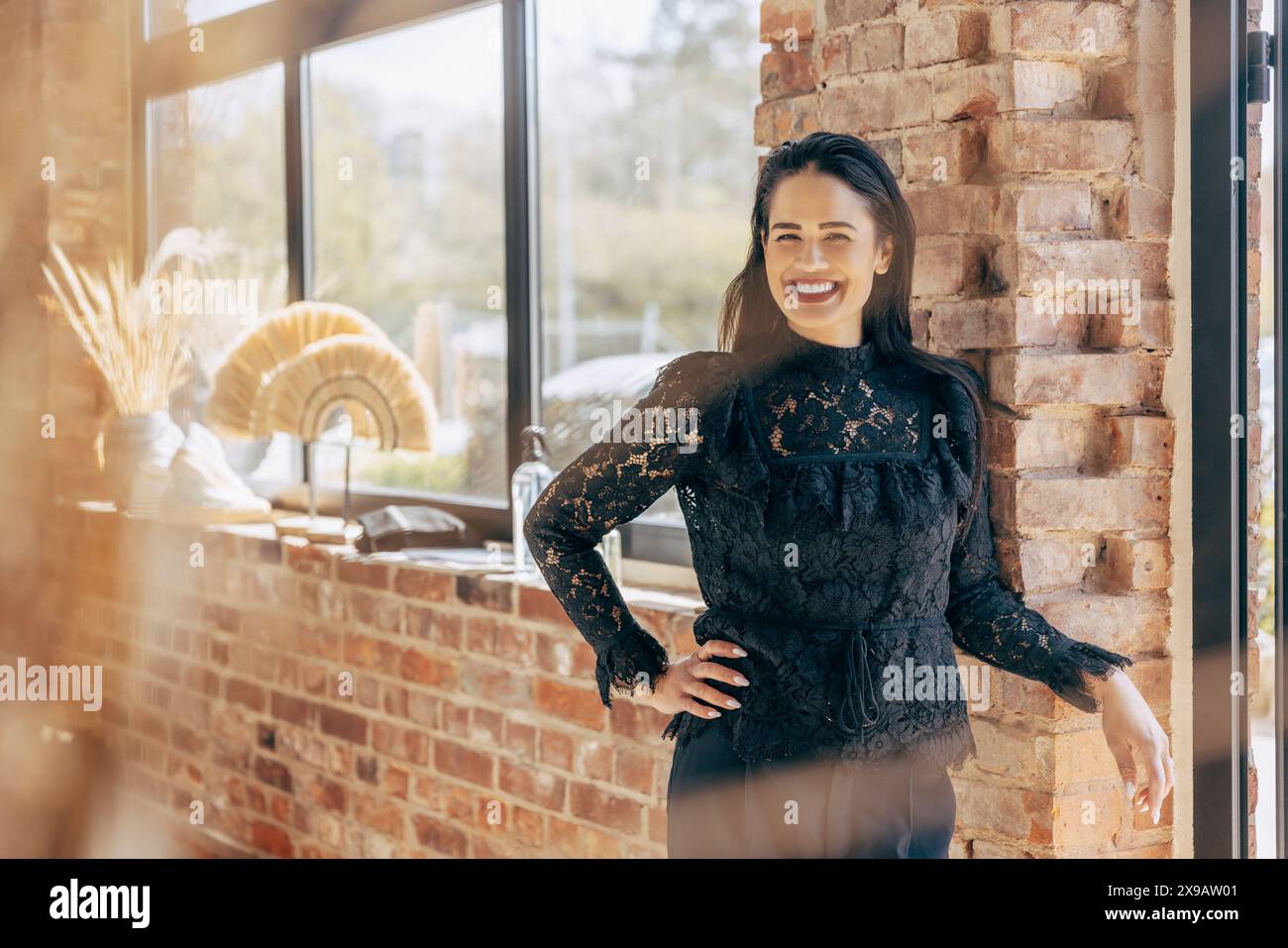 Portrait of smiling female owner with hand on hip leaning on brick wall ...