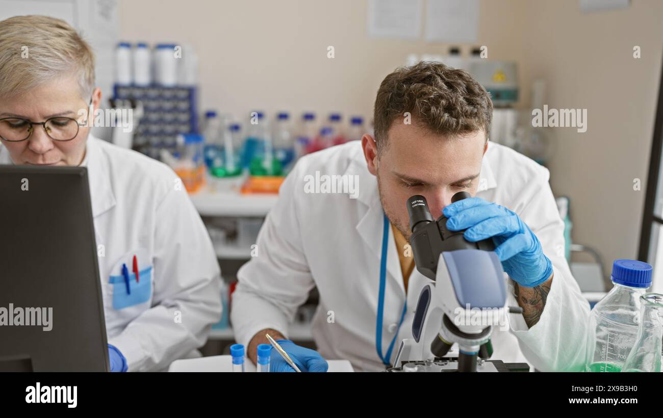A male scientist using a microscope beside a female colleague, both in ...