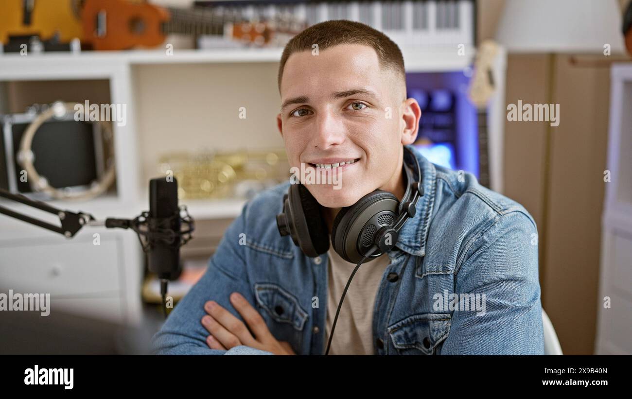 Handsome young man with headphones smiles confidently in a music studio setup, with a microphone ...
