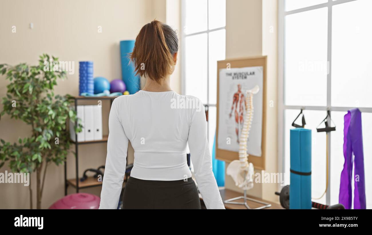 A young adult woman stands in a gym, facing away, surrounded by fitness ...