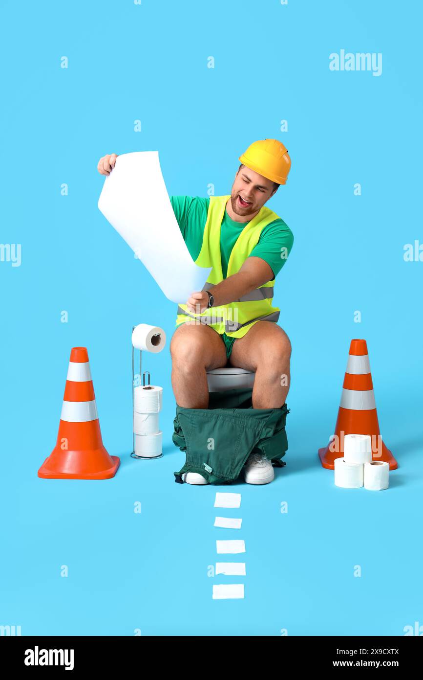 Male worker sitting on toilet bowl with plan and traffic cones against ...