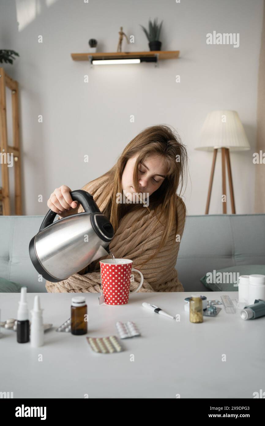 sick girl sitting on sofa in room holds kettle and pours boiling water ...