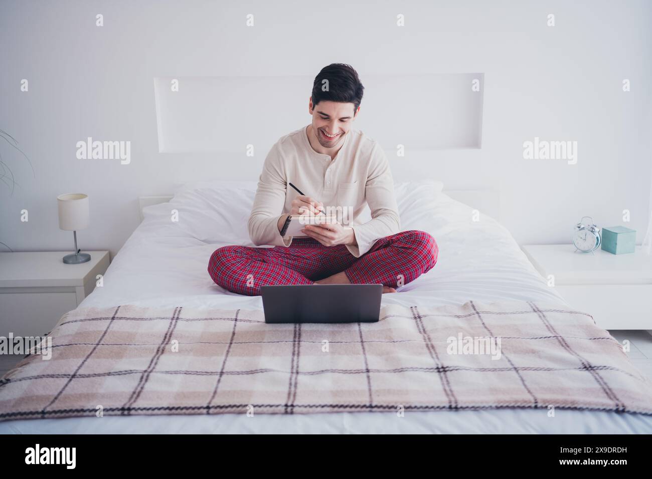 Photo of cheerful nice man sitting on bed writing notes weekly plan ...