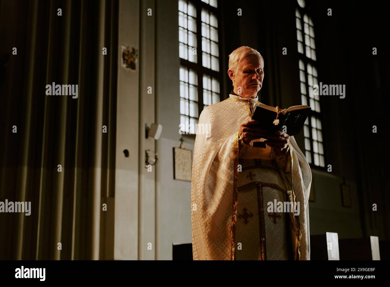 Medium long shot of senior Catholic priest wearing liturgical clothes standing indoors reading ...