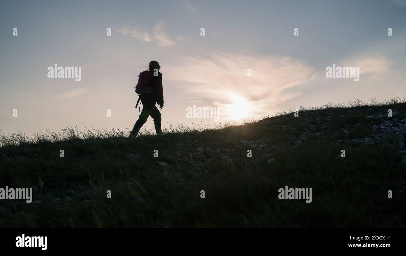 Female hiker silhouette with a backpack walking and following the trail uphill, enjoying the sky ...