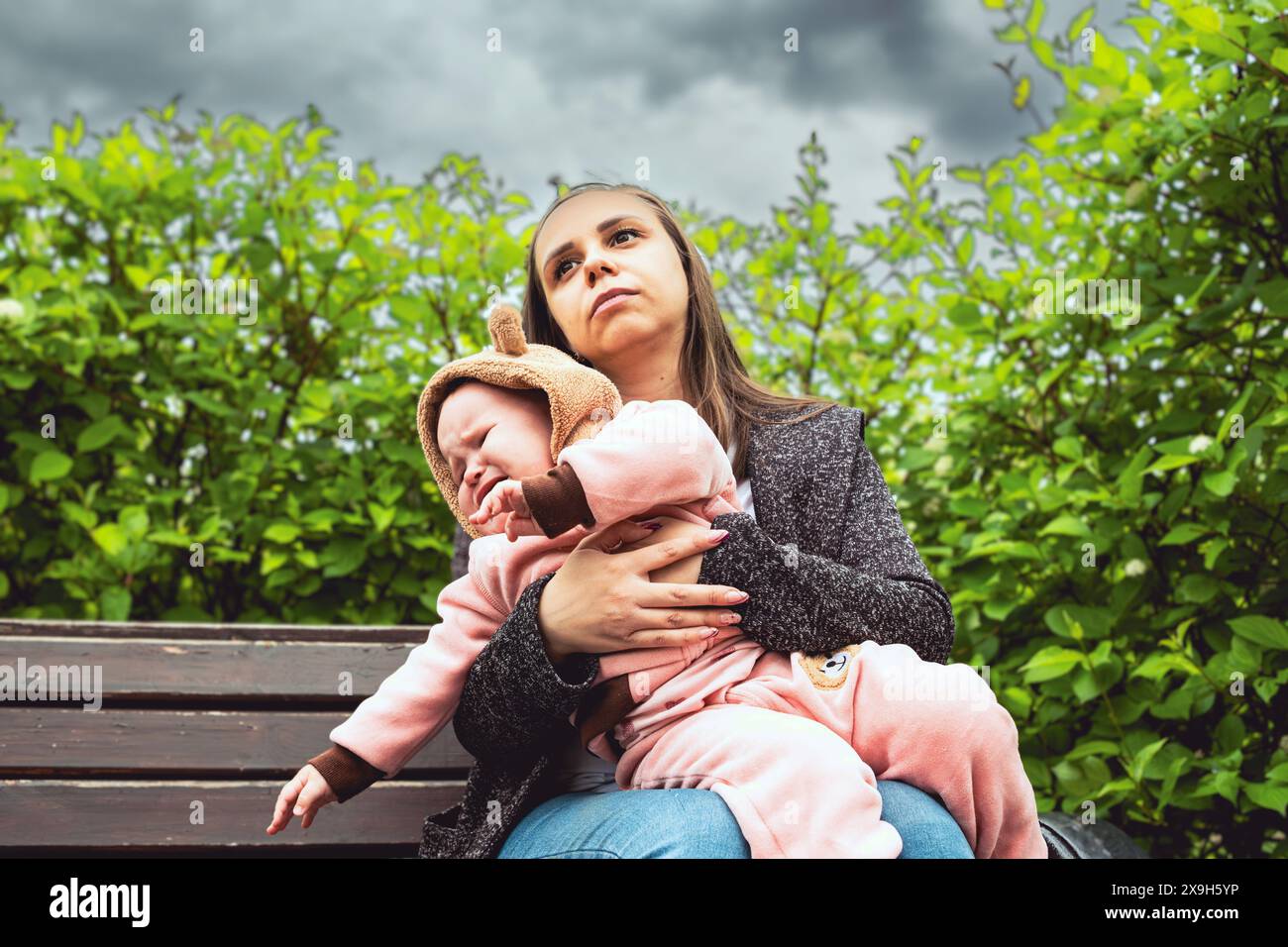 A tired mother sits on a park bench with her baby who is crying ...