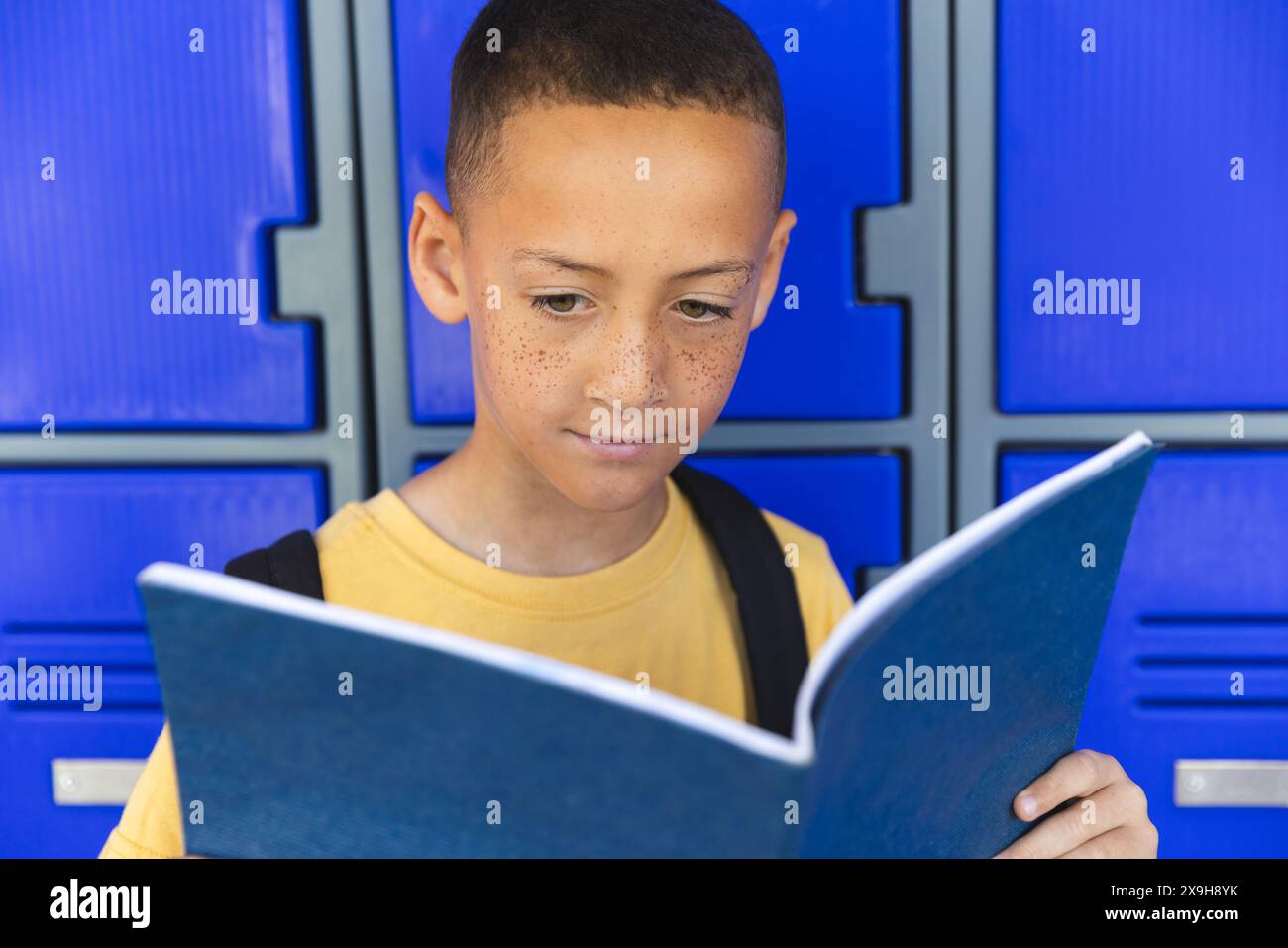 In a school, a young biracial boy stands before a blue locker ...