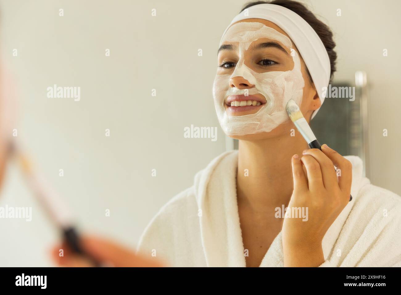 At home, young Caucasian woman applying face mask with brush, smiling ...