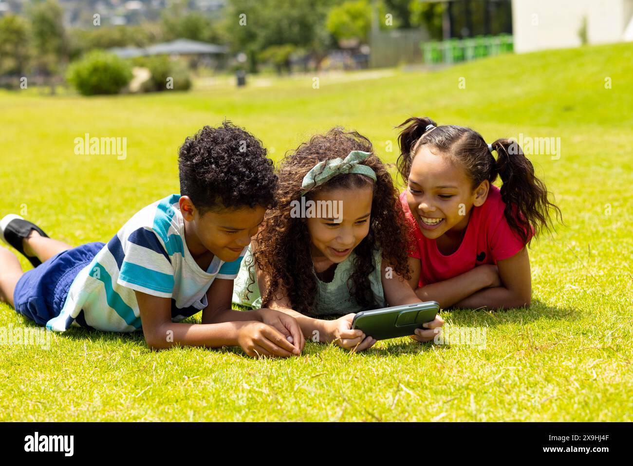 Three biracial children are lying on the grass, looking at a smartphone ...