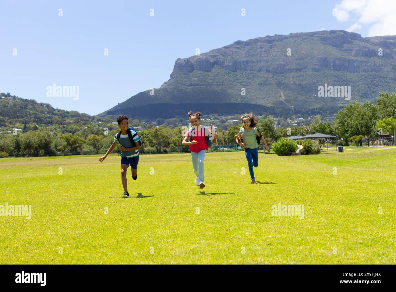 Three biracial children run across grassy field, mountains behind Stock ...