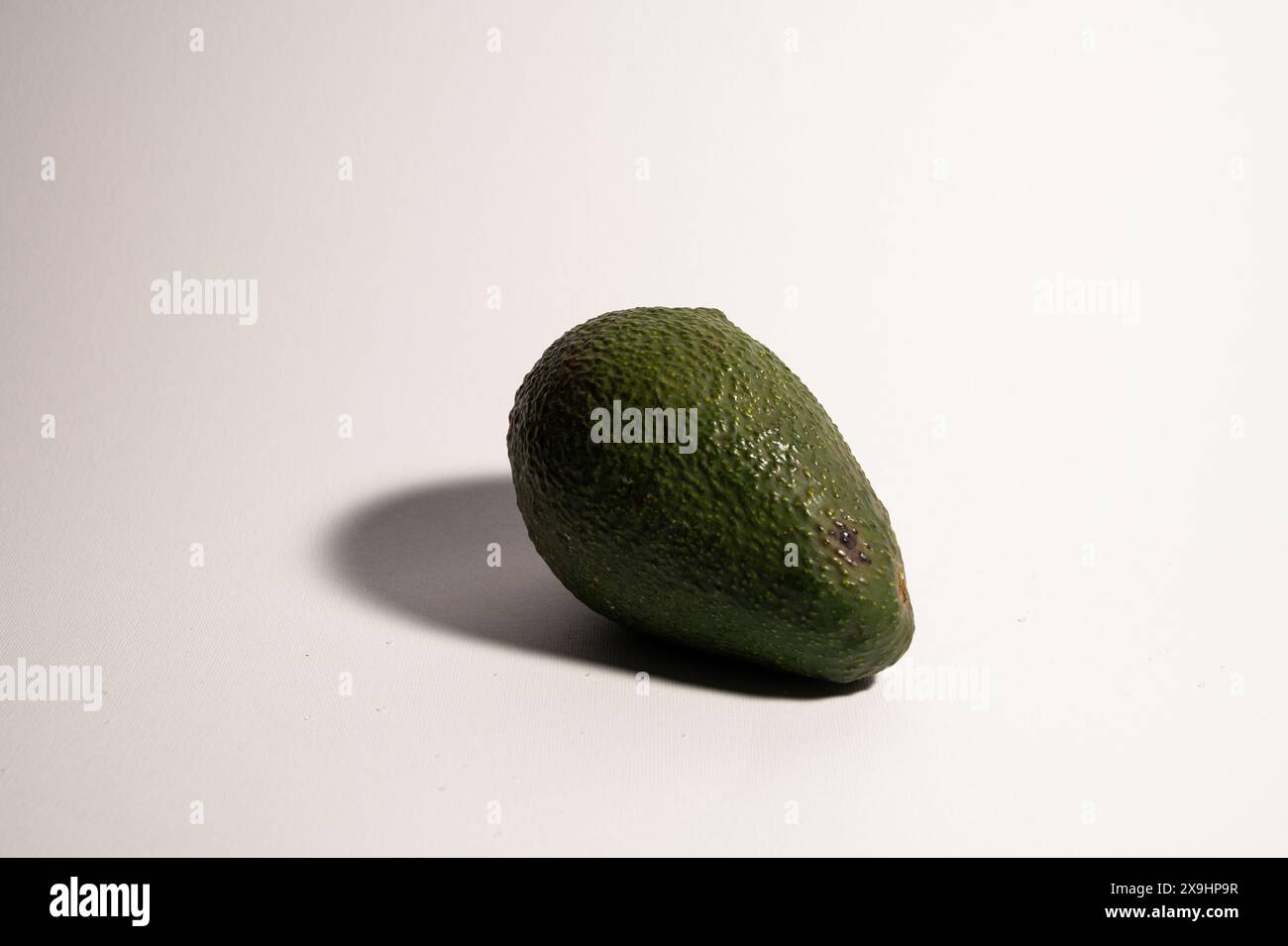 A green avocado sits on a white background. The avocado is unpeeled and ...