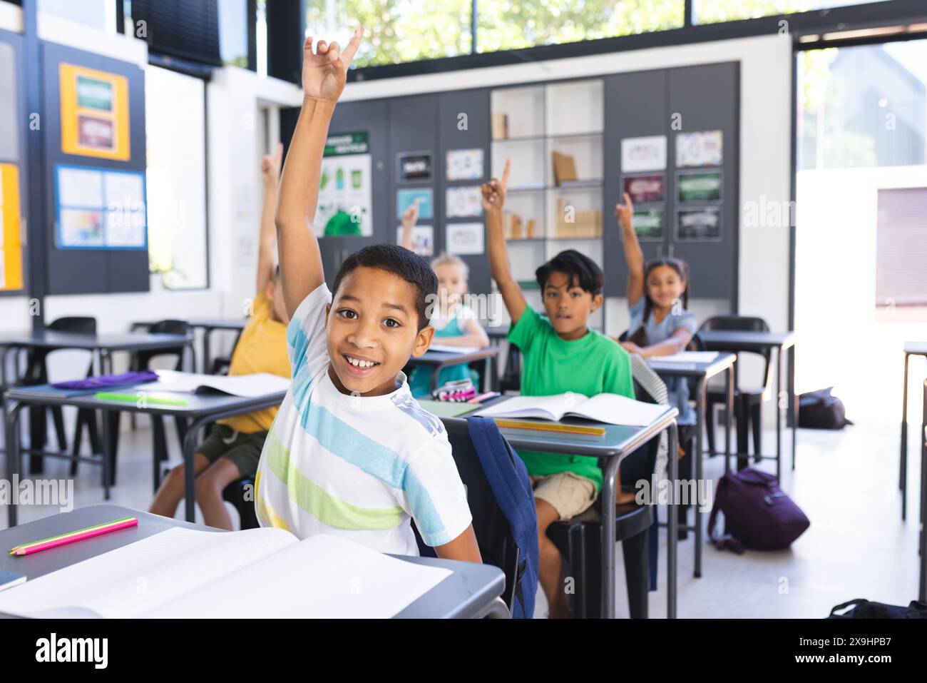 Biracial boy raises his hand eagerly in a classroom. Other children ...