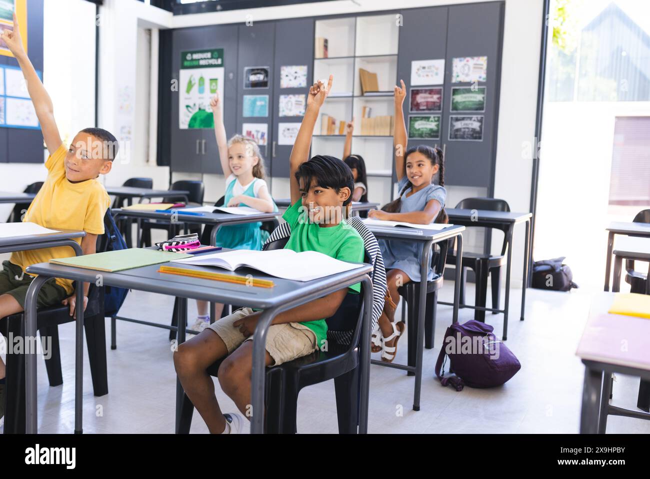Diverse group of children raising hands eagerly in a school classroom ...