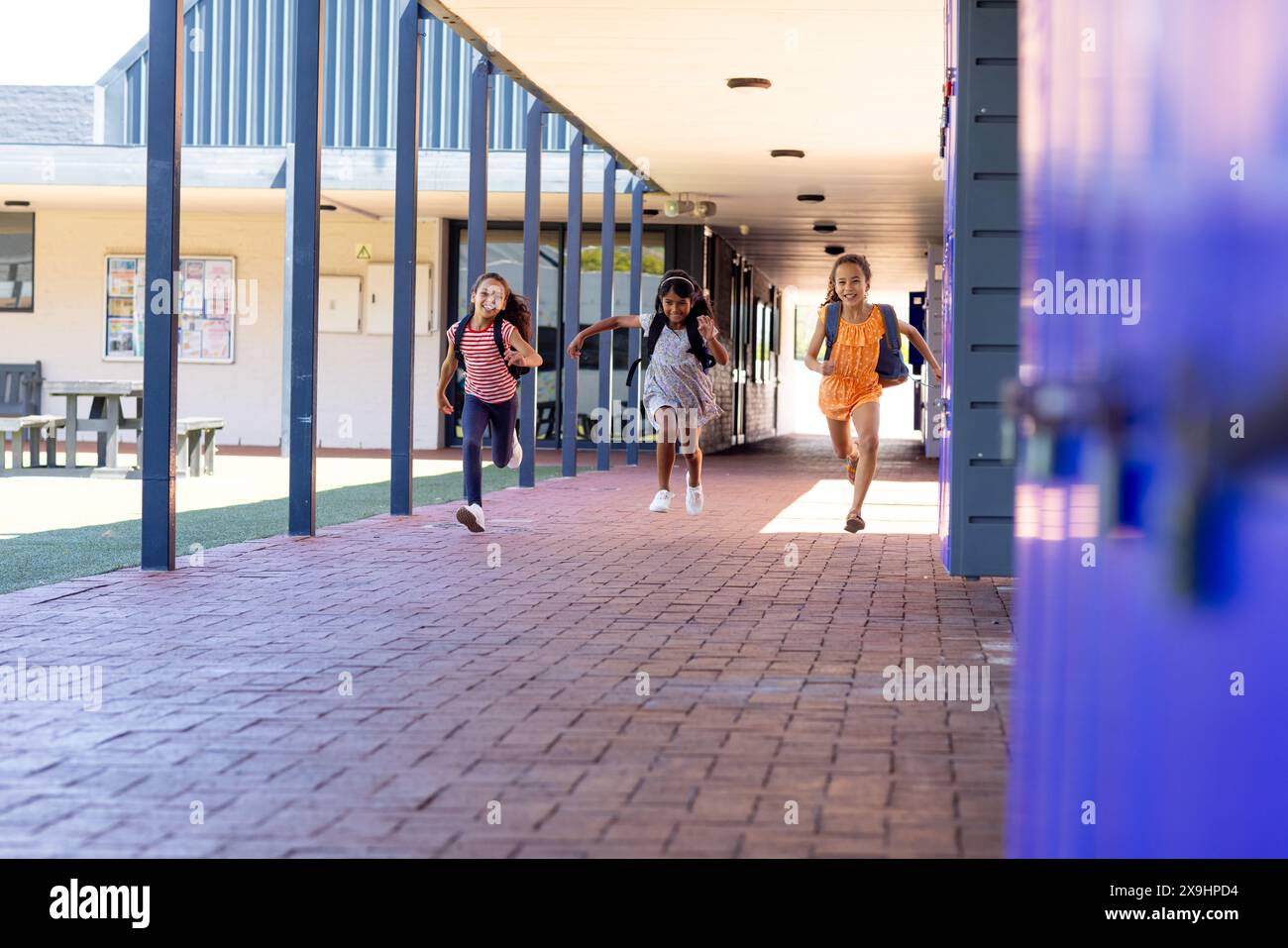 Three biracial girls are running joyfully down a school corridor with ...