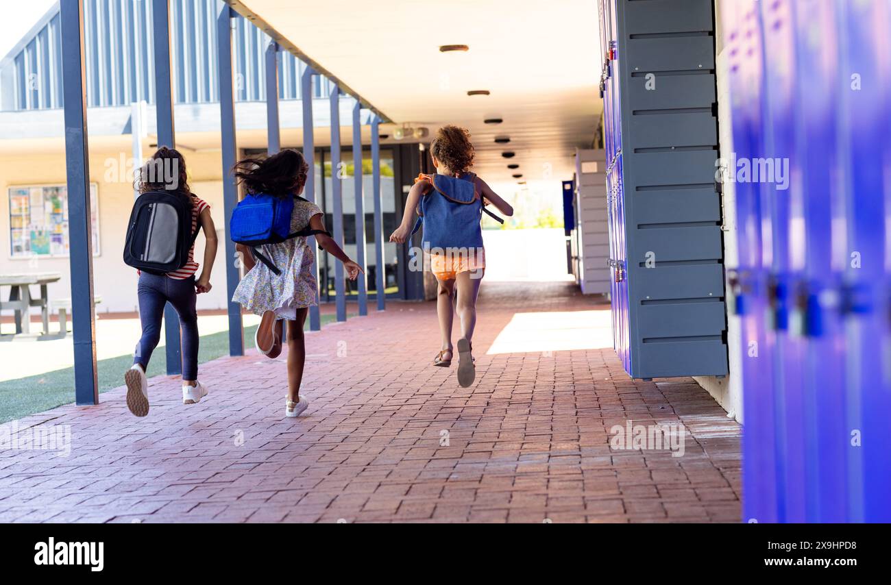 Three biracial girls are running through a school corridor, backpacks ...