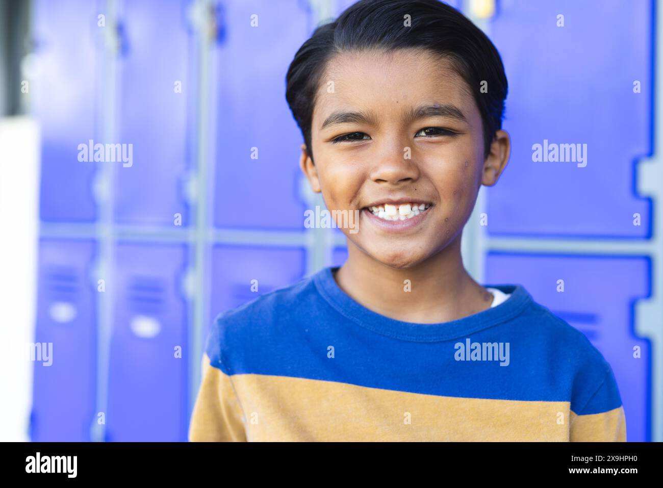 Biracial boy with a bright smile stands in front of blue lockers in ...