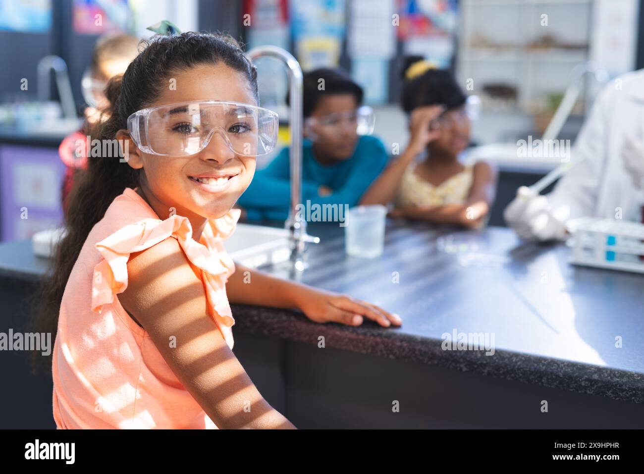 A biracial girl with safety goggles smiles in a science classroom at ...