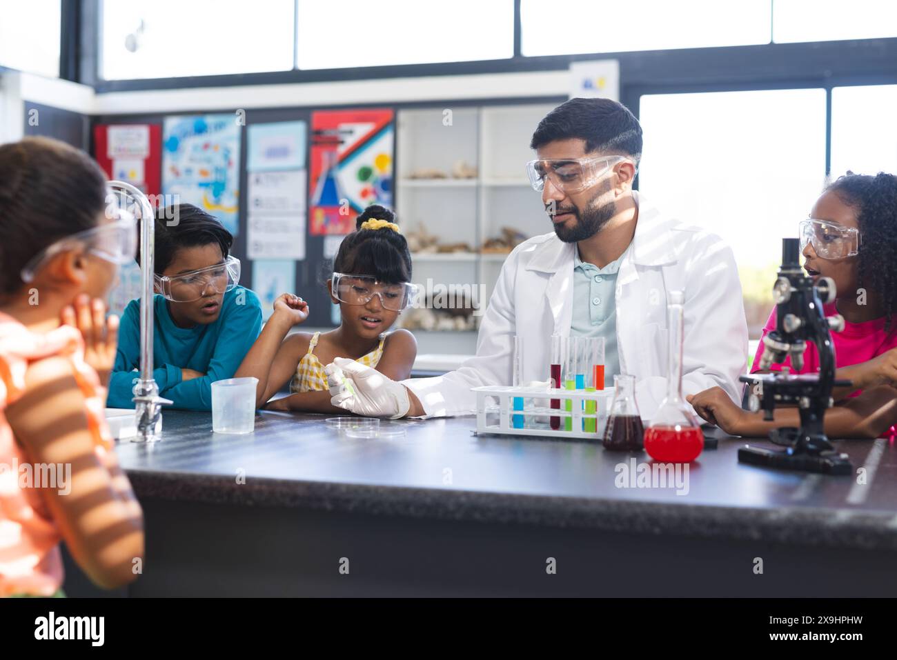 A young Asian male teacher shows experiment. Biracial kids in goggles ...