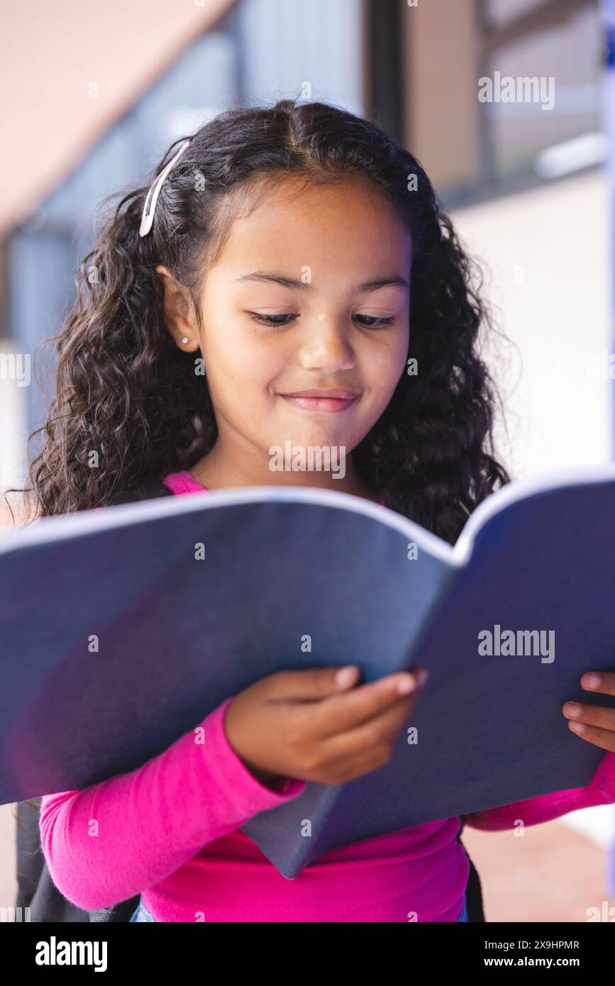 In school, young biracial girl holding a blue book, reading with ...