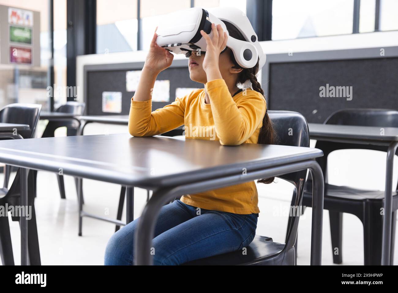 In school, young biracial girl wearing a VR headset sits at a table in ...