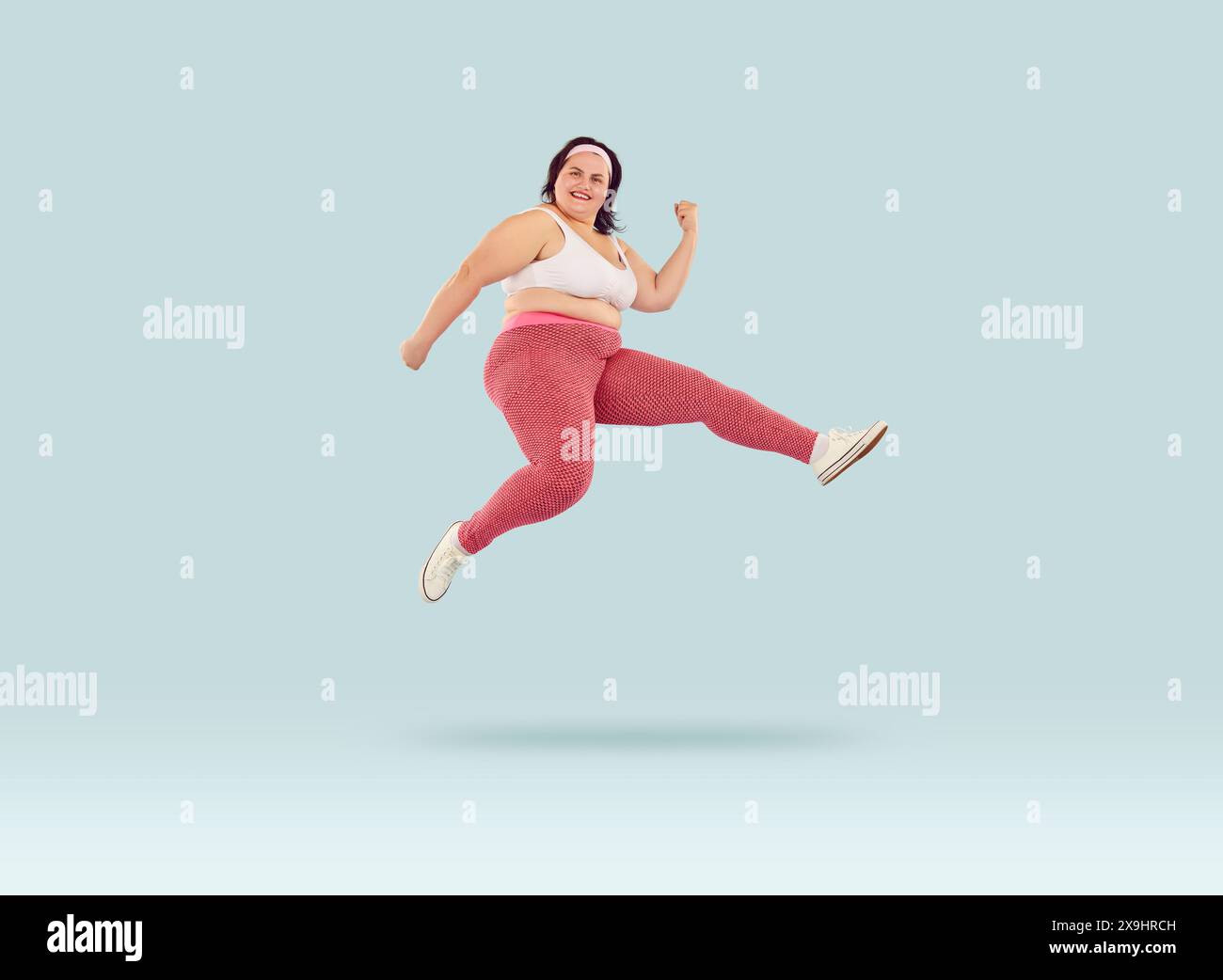 Fat happy woman wearing sportswear jumping isolated on studio blue ...